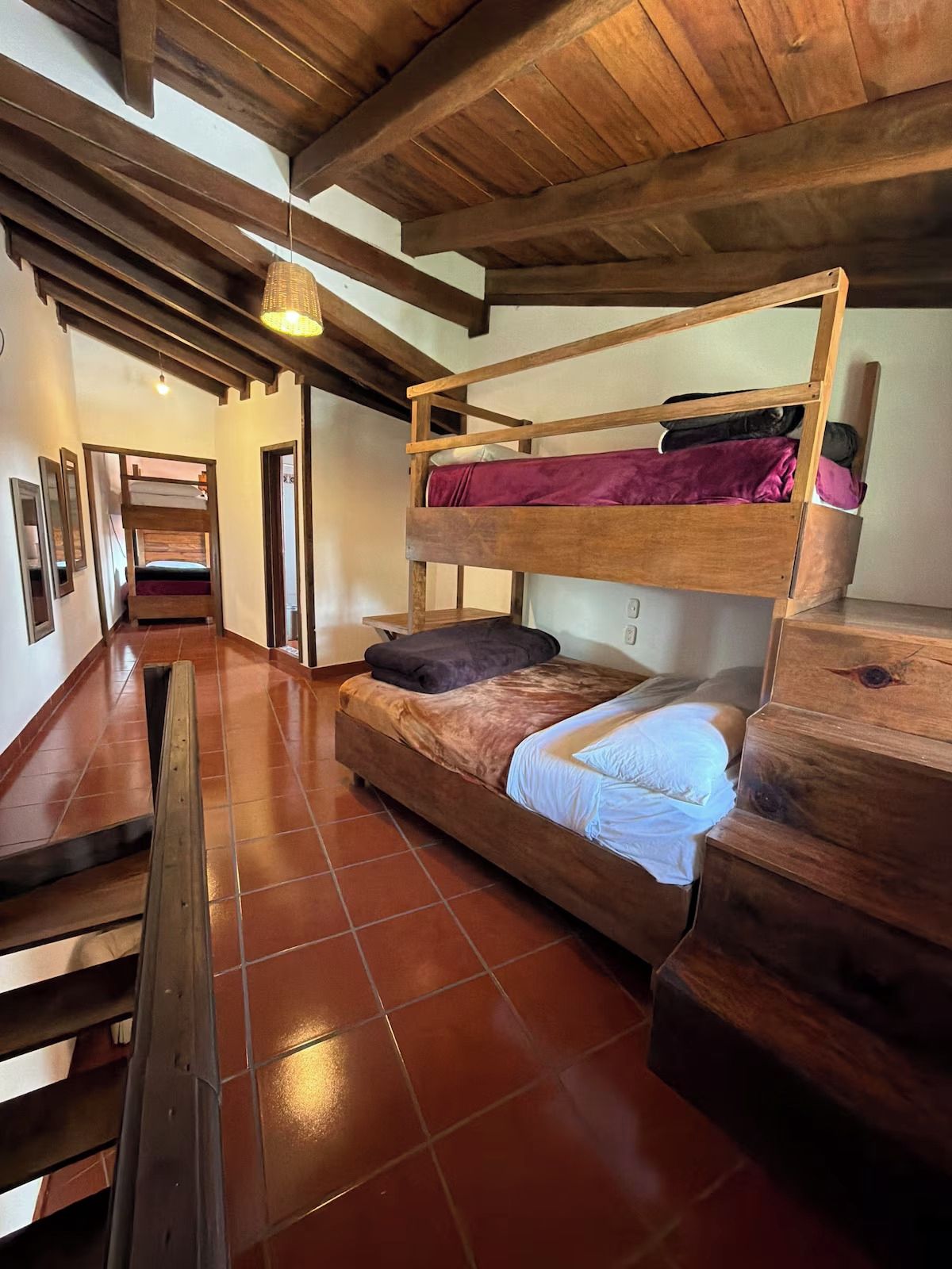 Quarto com beliches, vigas de madeira, um banco e luminária pendente.