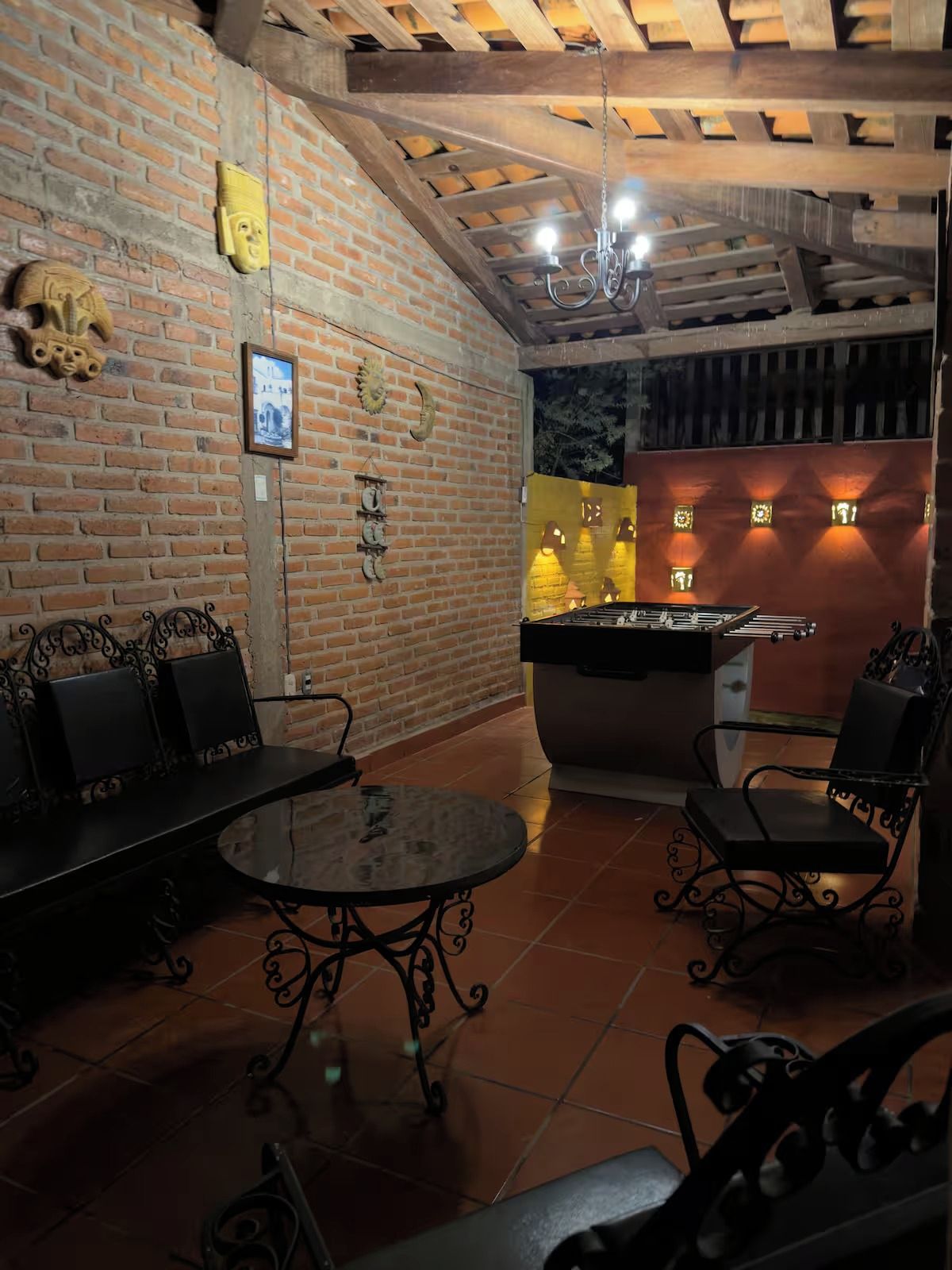 Um salão com paredes de tijolo, mesa de bilhar, duas cadeiras de metal preto, uma mesa redonda e teto com vigas aparentes.