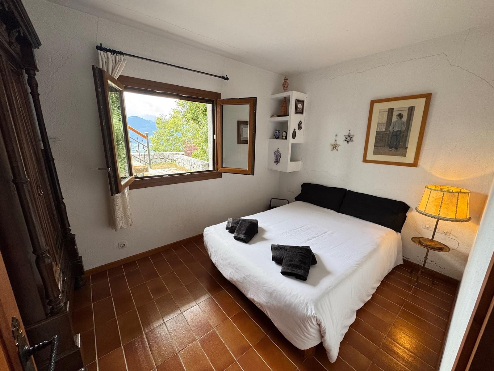Quarto com cama, janela, luminária e prateleira com objetos decorativos.