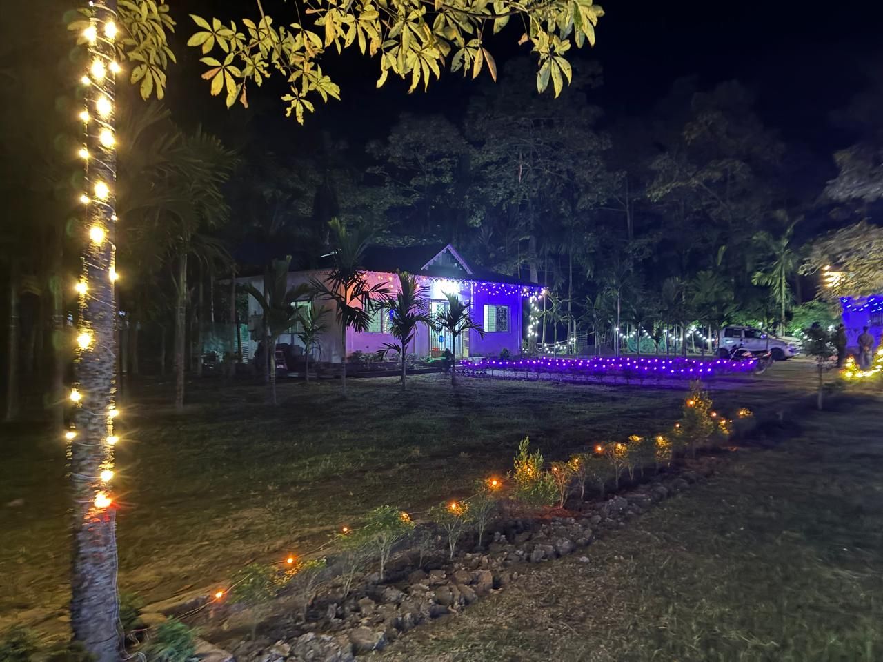 Área externa iluminada à noite por luzes de corda, com um pequeno edifício e um caminho.