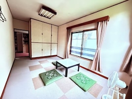 Quarto com armário de porta deslizante, uma mesa, dois tapetes verdes, um ventilador e uma janela com cortinas.