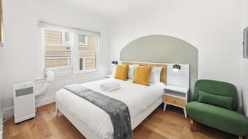Quarto de hotel com cama, mesa de cabeceira, cadeira verde, janela com persianas e radiador.