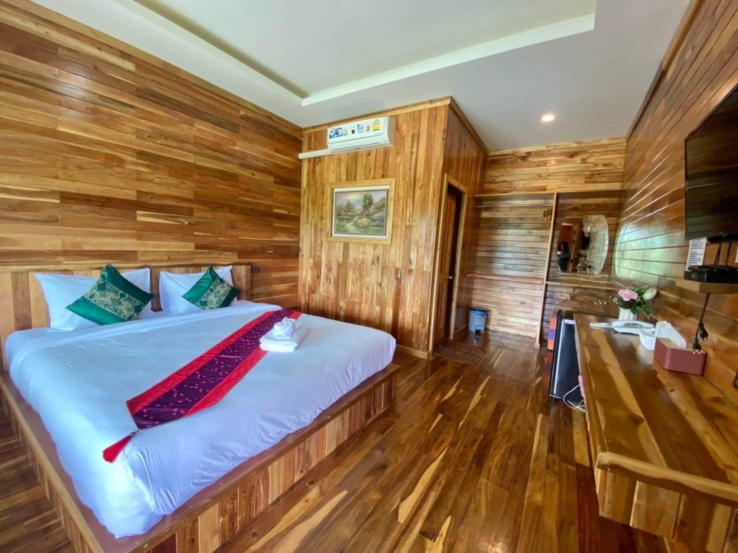 Quarto de hotel com painel de parede em madeira, cama com roupa de cama branca e almofada decorativa vermelha, pequena escrivaninha com espelho e ar condicionado instalado no teto.