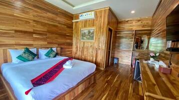 Quarto de hotel com painel de parede em madeira, cama com roupa de cama branca e almofada decorativa vermelha, pequena escrivaninha com espelho e ar condicionado instalado no teto.