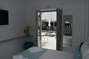 Quarto de hotel com cama, banheiro com pia e espelho, e porta aberta para área da piscina com cadeiras de descanso.