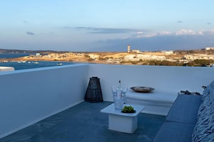 Terraço no telhado com vista para o mar e edifícios ao longe, uma mesa com garrafa e copos, e um sofá de vime.