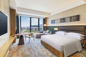 Quarto de hotel com uma cama grande, escrivaninha com cadeira, sofá e uma janela ampla com vista para a cidade.