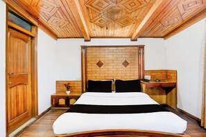 Quarto de hotel com teto de madeira, cabeceira estampada em tijolos, cama com roupa de cama branca, mesa de cabeceira e escrivaninha com telefone.