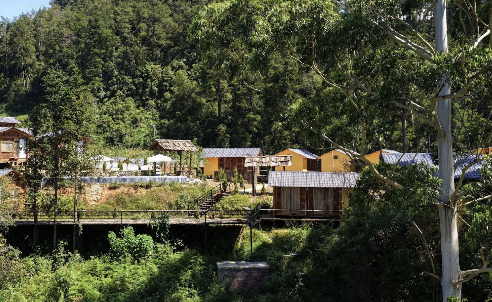 Paisagem rural com uma ponte de madeira, um edifício amarelo e uma colina arborizada.