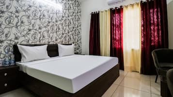 Quarto de hotel com cama, mesa de cabeceira, cadeira e janela com cortinas.