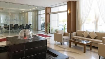 Um saguão de hotel com balcão de recepção, uma fonte, área de estar e janelas amplas.
