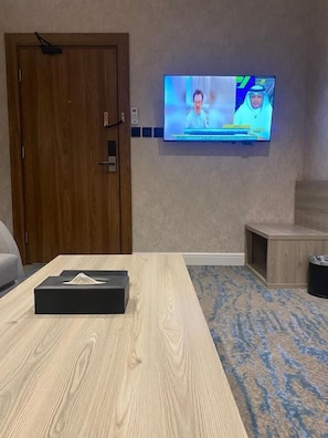 Um quarto com porta de madeira, televisão fixada na parede, uma pequena mesa de madeira e um tapete estampado.