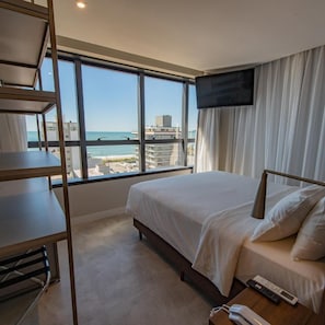Quarto de hotel com cama, escrivaninha, televisão e vista para a praia e edifícios.