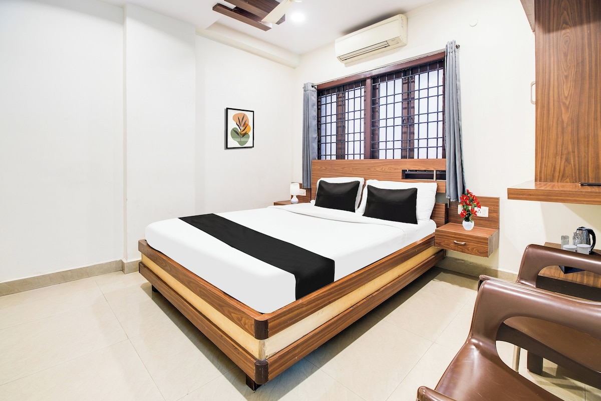 Quarto de hotel com cama de madeira, mesa de cabeceira, escrivaninha com cadeira e janela com grades.