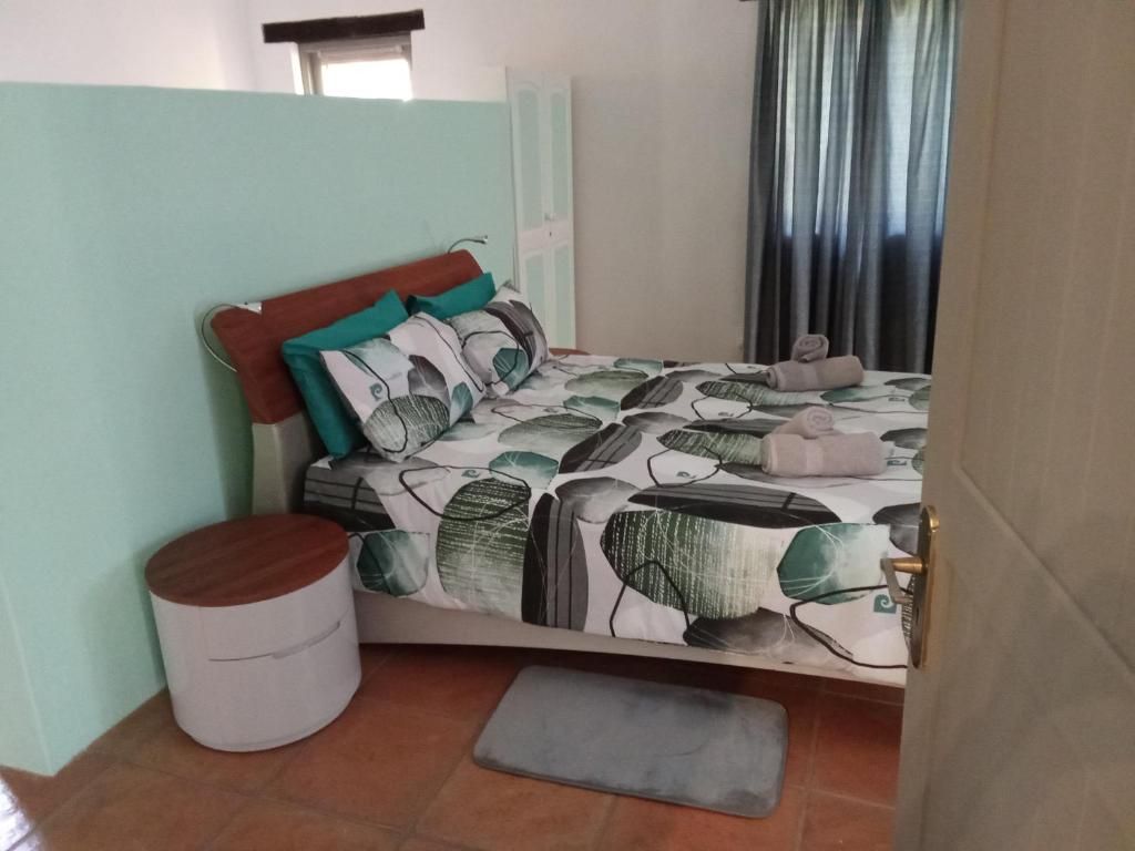 Uma cama com colcha estampada com folhagens, uma mesinha de cabeceira e um tapete pequeno sobre piso de azulejos.
