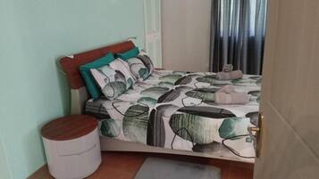 Uma cama com colcha estampada com folhagens, uma mesinha de cabeceira e um tapete pequeno sobre piso de azulejos.