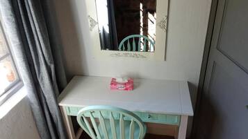 Quarto com escrivaninha branca, cadeira azul clara, espelho com detalhes decorativos e janela com cortinas.