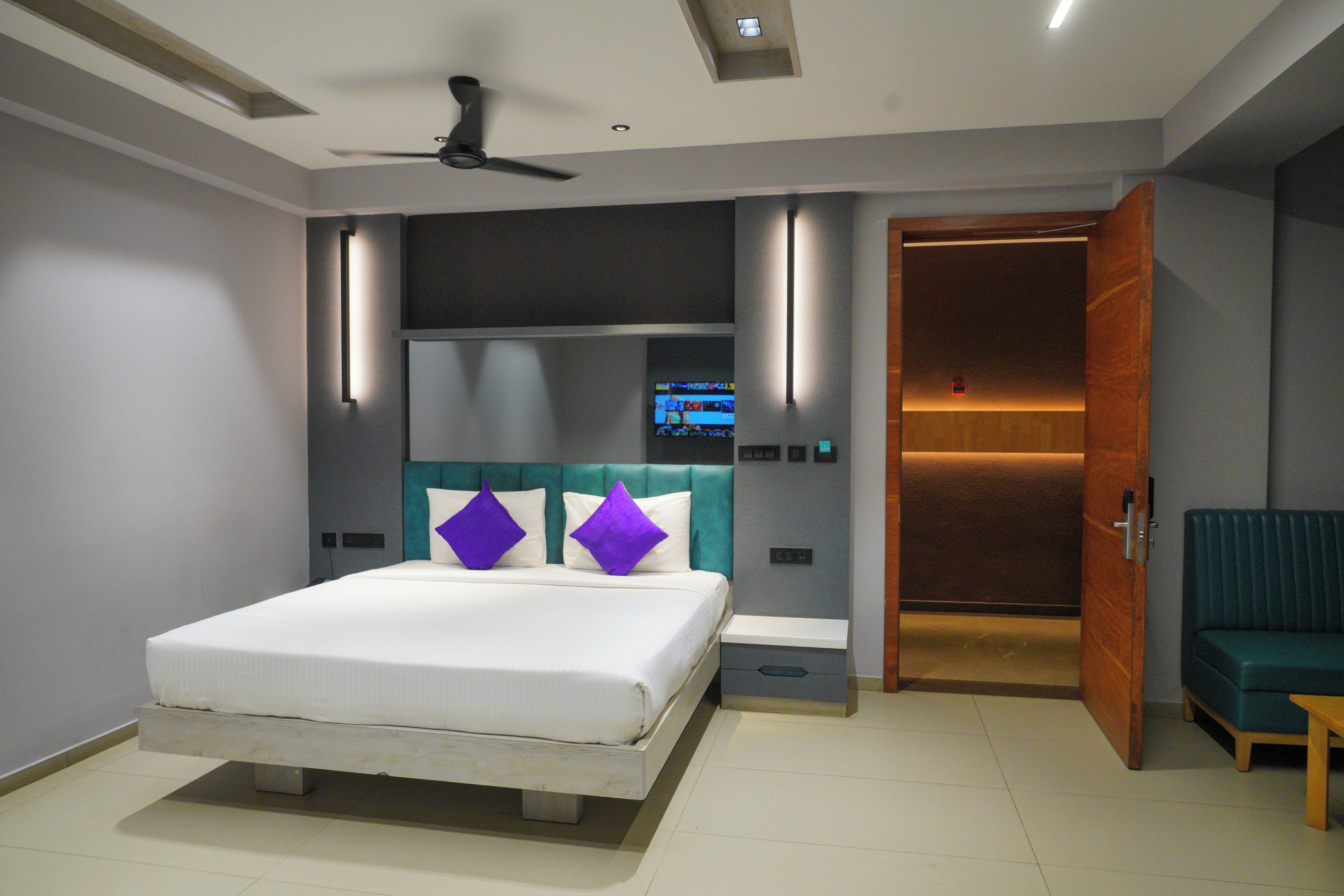 Quarto de hotel moderno com cama, televisão, ventoinha no teto e porta de madeira.