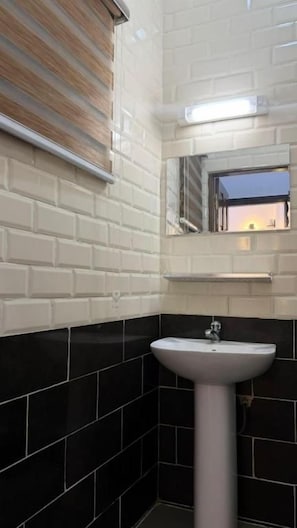 Casa de banho com lavatório pedestal branco, paredes revestidas a mosaico preto, janela com persianas e espelho sobre o lavatório.