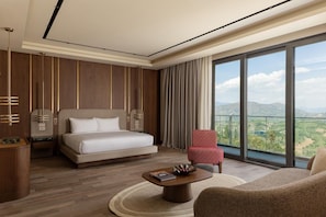 Quarto de hotel com uma cama grande, área de estar com mesinha de café, uma poltrona vermelha com padrão e vista para a paisagem através de amplas janelas.