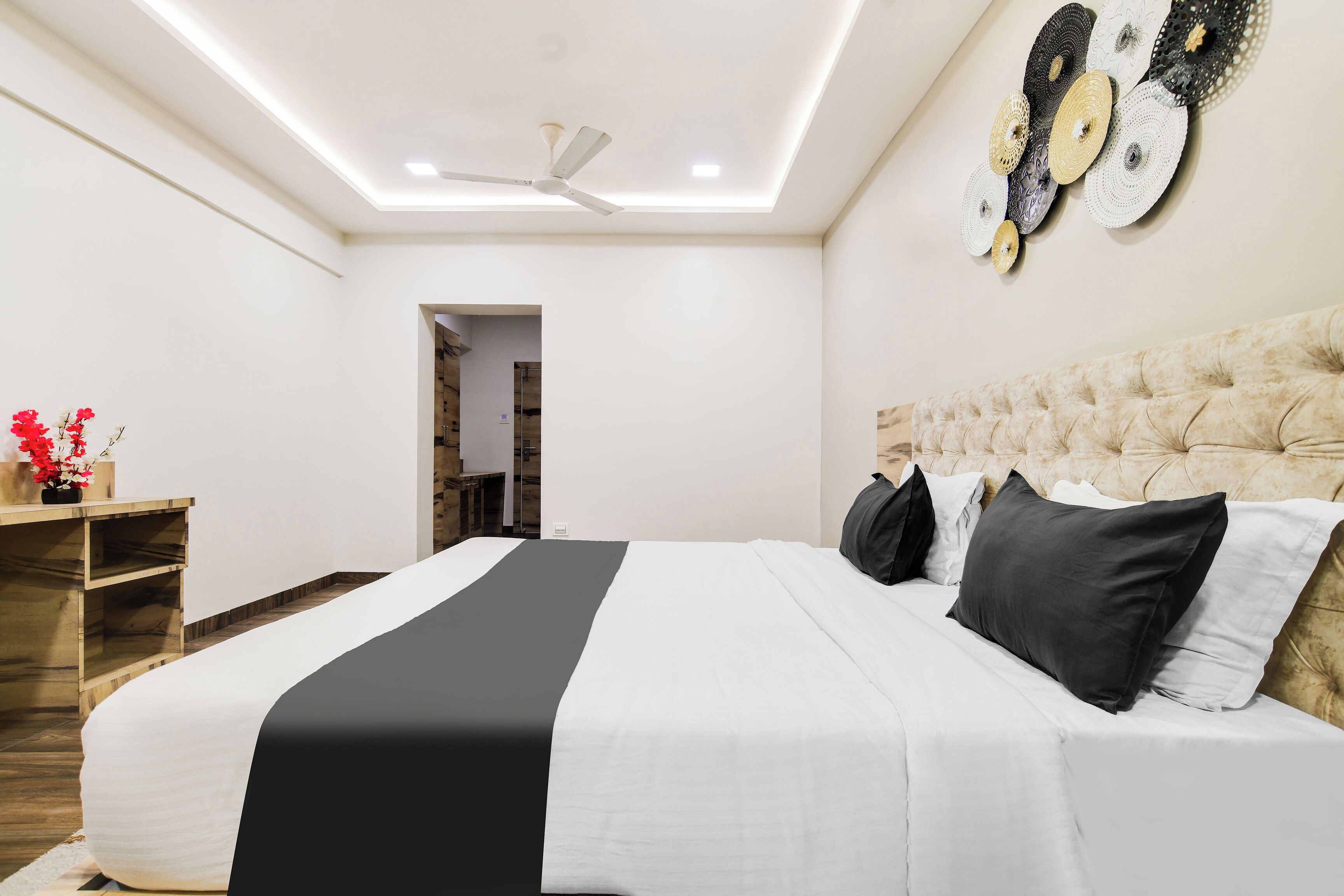 Quarto com cama, ventoinha no teto, objeto decorativo na parede e prateleira de madeira com flores.