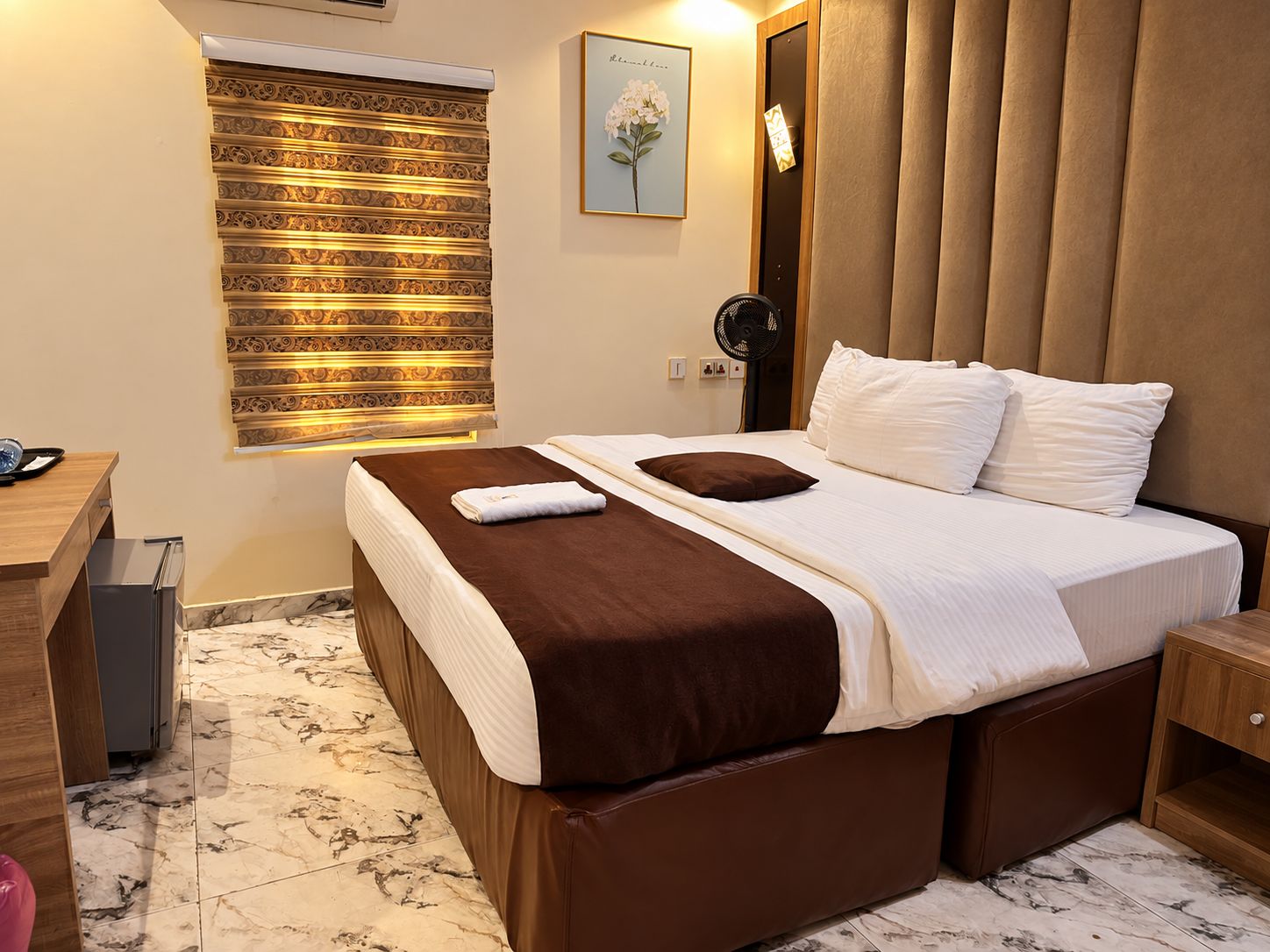 Quarto de hotel com cama, mesa de cabeceira e ventoinha na parede.
