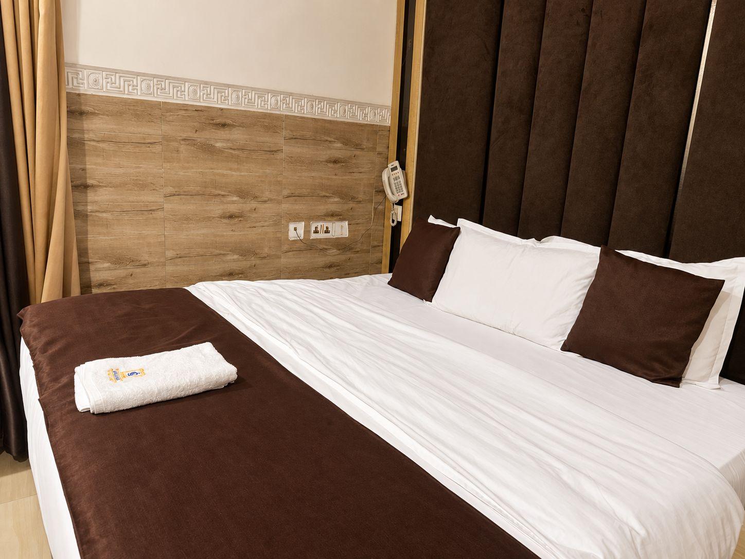 Quarto de hotel com cama, roupa de cama marrom e branca, telefone e uma moldura decorativa na parede.