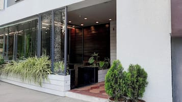 Entrada de prédio com porta de vidro, plantas em vasos e um pequeno degrau para o interior.