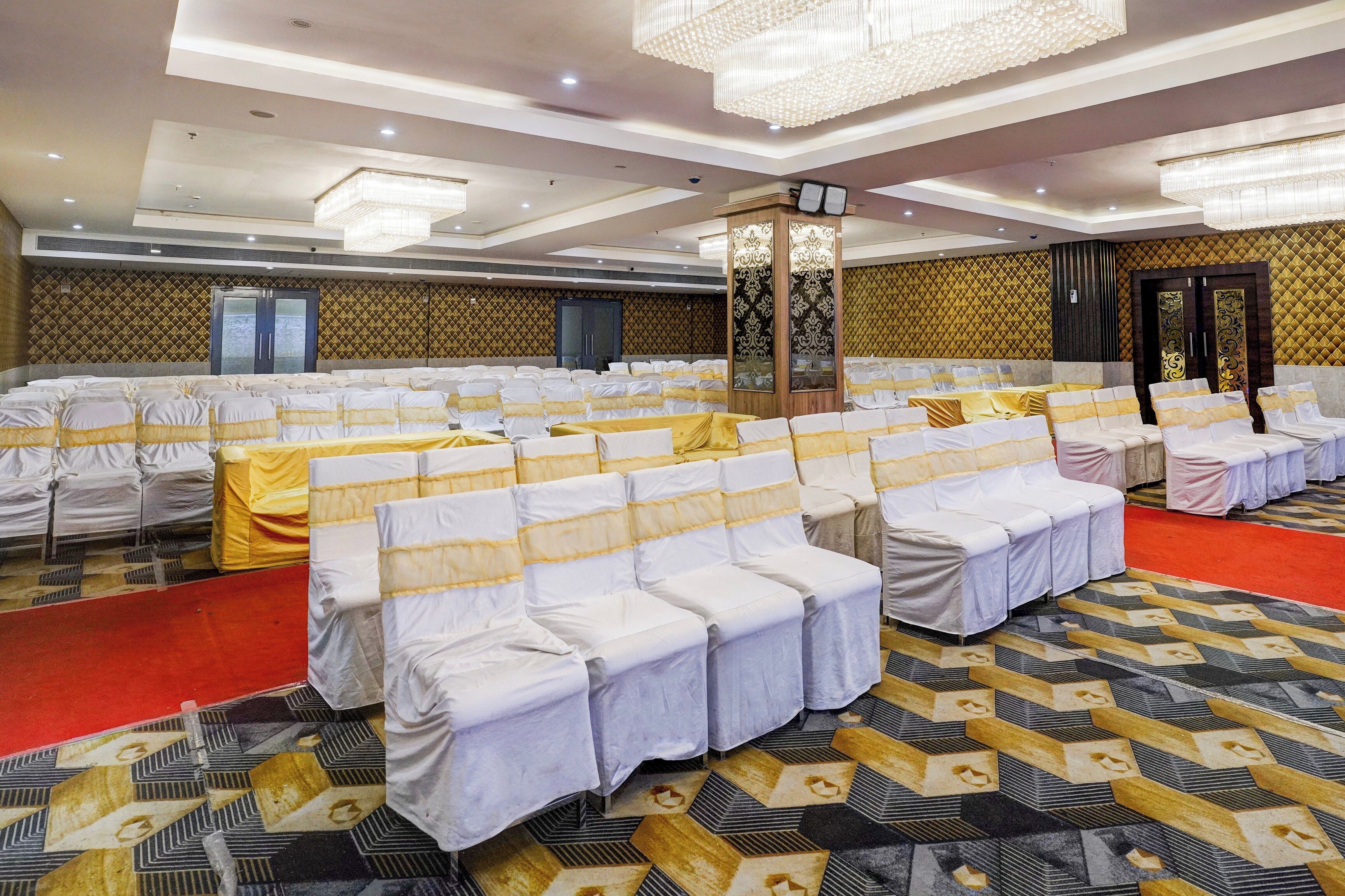 Sala de conferências com fileiras de cadeiras brancas, uma coluna central e carpete estampado.