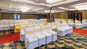 Sala de conferências com fileiras de cadeiras brancas, uma coluna central e carpete estampado.