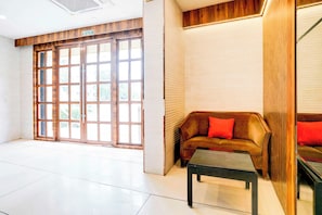Um saguão de hotel com uma grande porta de vidro emoldurada em madeira, um sofá com almofadas vermelhas e uma pequena mesa.