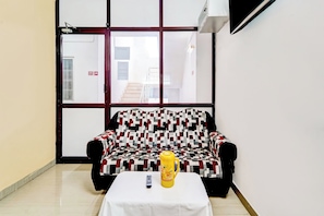 Um quarto com um sofá estampado, uma mesa pequena com um controle remoto e uma caneca amarela, e uma janela com o aviso de saída de incêndio.