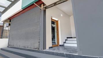 Porta de persiana fechada, um lance de escadas que levam a uma porta de madeira e uma pequena área externa com cerca de metal.