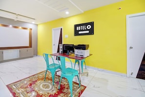 Quarto de hotel com parede amarela, placa indicando que é um hotel de serviço corporativo, escrivaninha com impressora, duas cadeiras turquesa e tapete estampado.