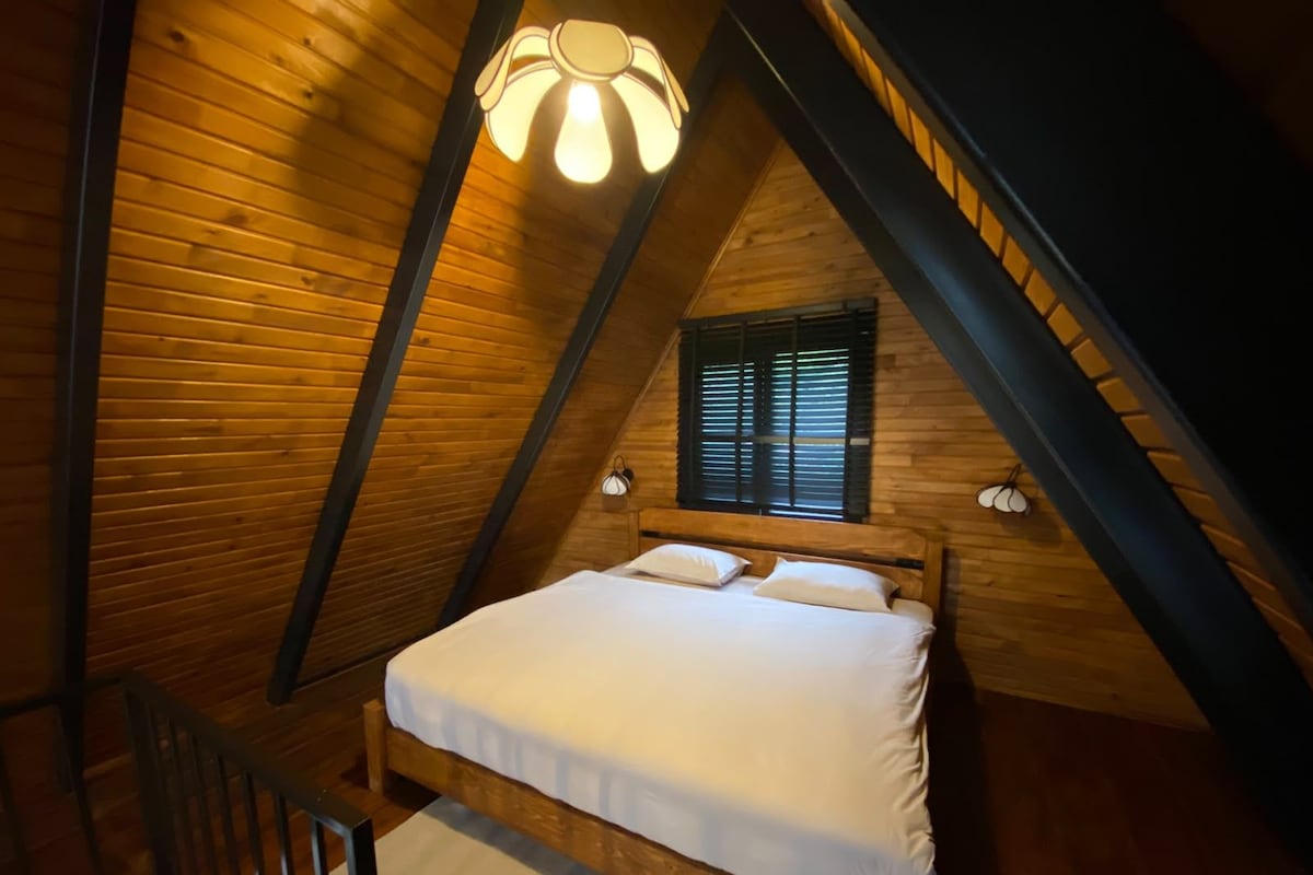 Quarto com teto de madeira inclinado, cama com roupa de cama branca e duas luminárias fixadas na parede.