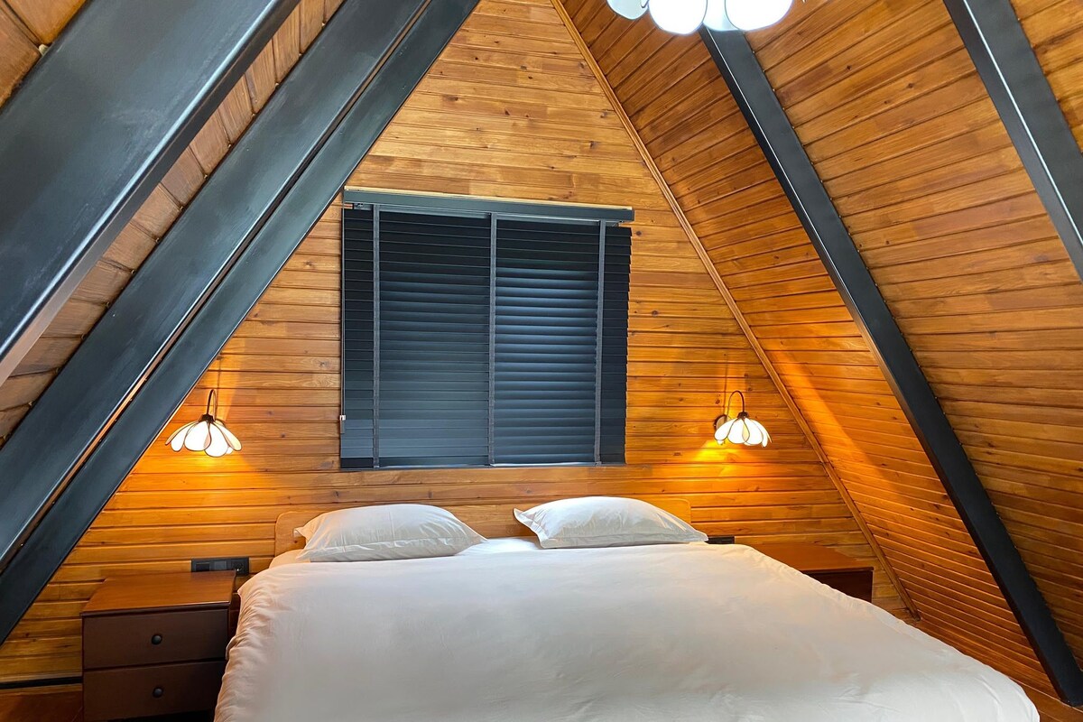 Quarto com teto inclinado, uma cama, duas mesas de cabeceira e uma janela com persianas.