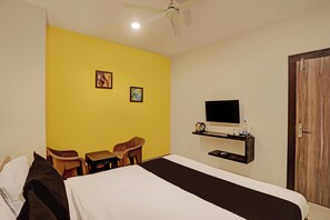 Quarto de hotel com cama, escrivaninha, duas cadeiras, televisão e porta.