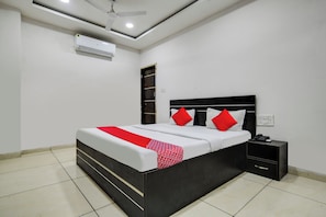 Quarto moderno com cabeceira preta, colcha branca e almofadas vermelhas. Há uma mesa de cabeceira com telefone e tomada.