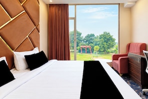 Quarto de hotel com uma cama grande, uma poltrona vermelha, cômoda de madeira e vista para um jardim.