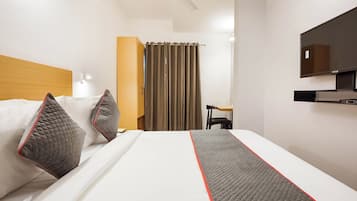Quarto de hotel com cama, almofadas, televisão, secretária e cortina.