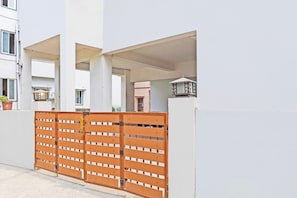 Uma entrada protegida por grades, com cerca de madeira, parede branca e um edifício ao fundo.