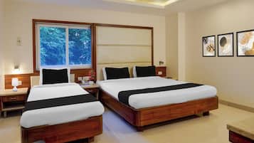 Quarto de hotel com duas camas, uma janela grande com vista para árvores, uma mesa de cabeceira com abajur e uma parede com obras de arte emolduradas.