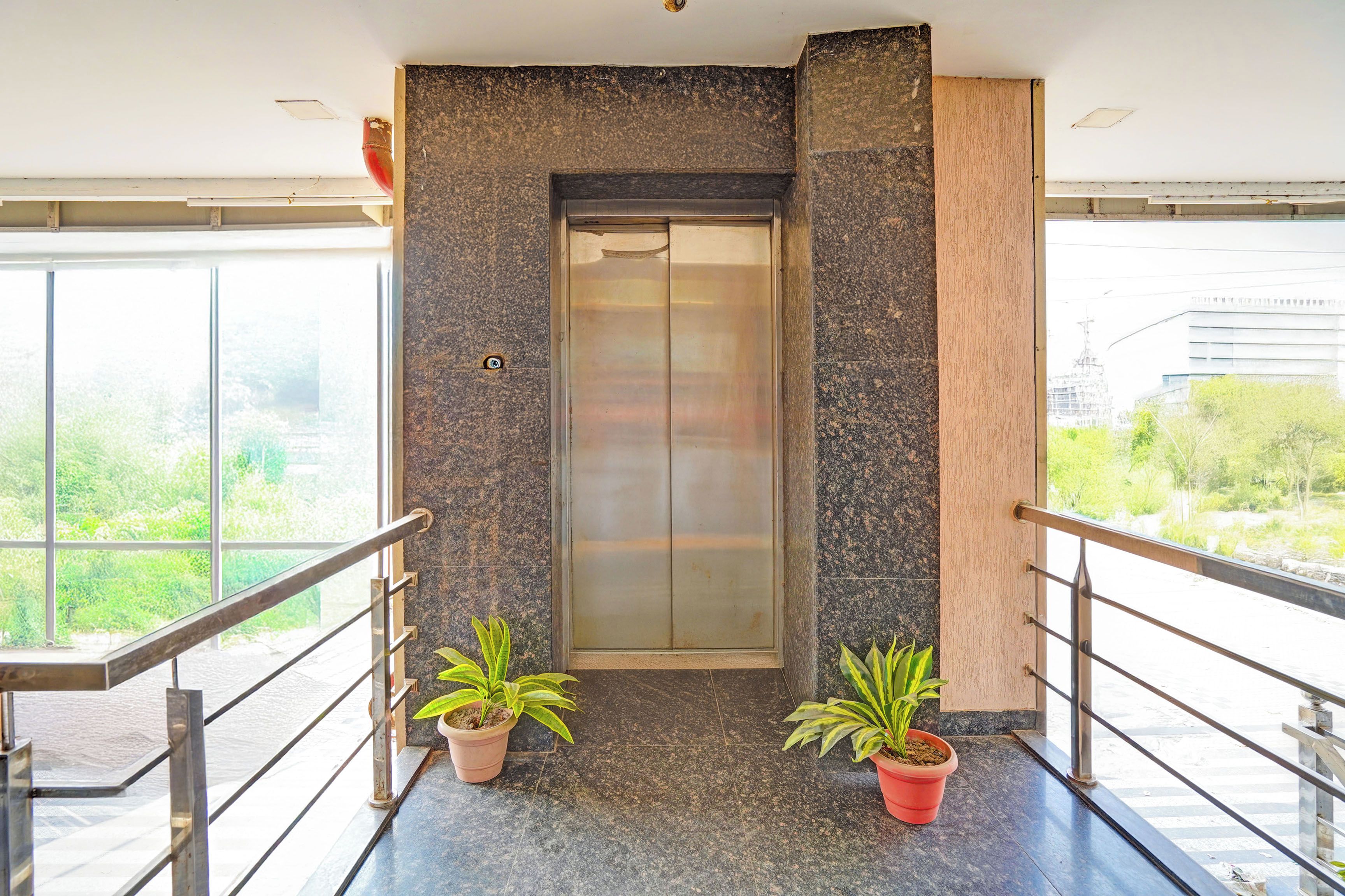 Entrada de elevador com porta de vidro, com plantas em vasos de cada lado, que dá para uma varanda com guarda-corpo de metal.