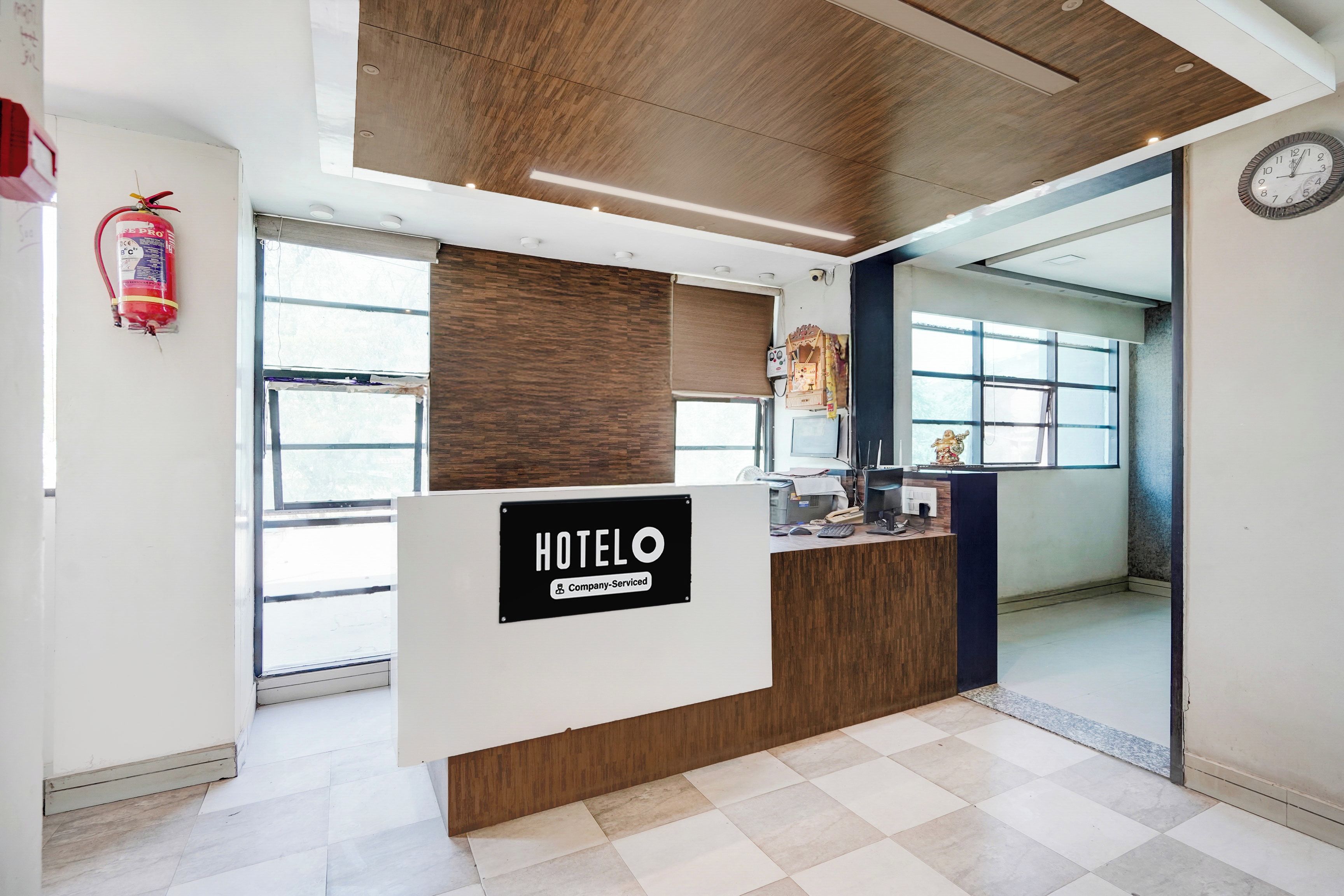 Balcão de recepção de hotel com uma placa 'HOTELO Company-Serviced', janelas amplas e teto com painéis de madeira.