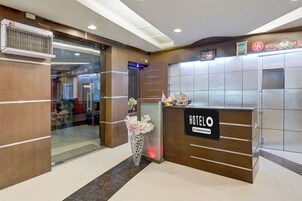 Balcão de recepção de hotel com sinalização indicando serviço empresarial. Entrada por porta de vidro, área de recepção com um vaso de flores e relógio digital mostrando a hora.
