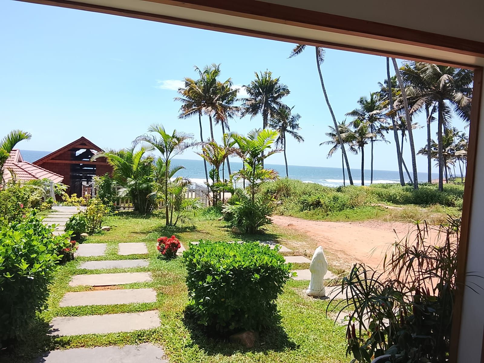 Caminho que leva a um edifício com telhado vermelho, cercado por vegetação exuberante e palmeiras, com vista para uma praia.