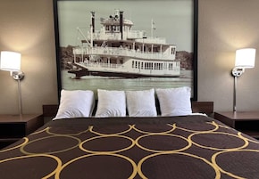 Quarto de hotel com cama, duas luminárias de cabeceira, um quadro de um barco na parede e um edredom estampado.