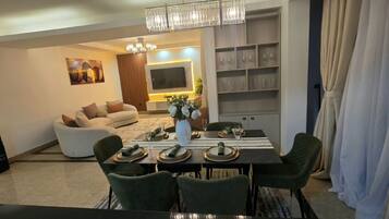 Uma sala de jantar com mesa posta para quatro pessoas, um sofá, uma TV e uma estante de livros.
