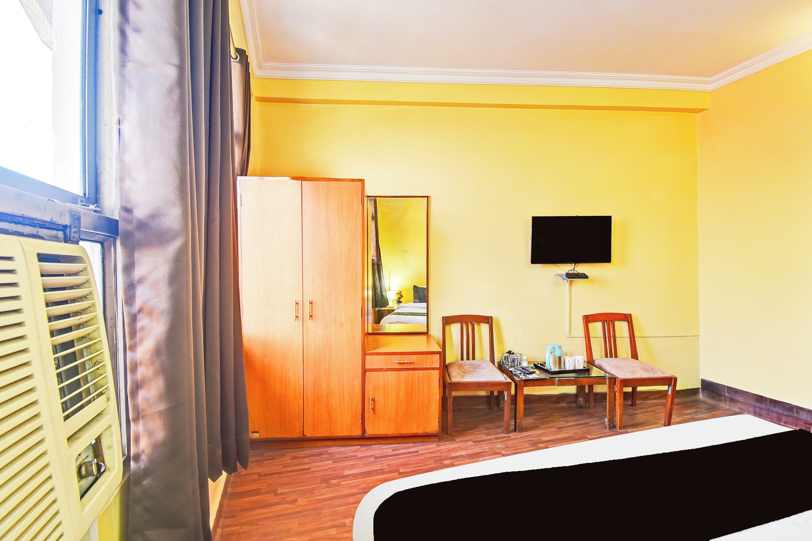 Quarto de hotel com cama, escrivaninha, cadeira, guarda-roupa, televisão e janela com cortinas.