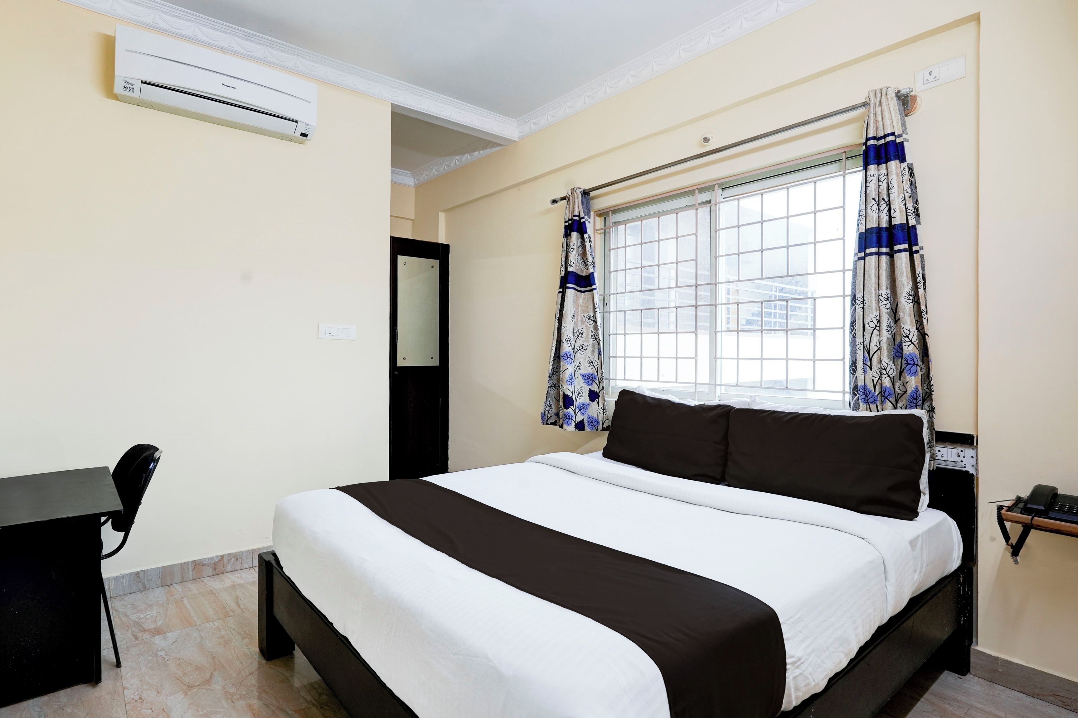 Quarto de hotel com cama, escrivaninha, ar-condicionado, janela com cortinas e porta.